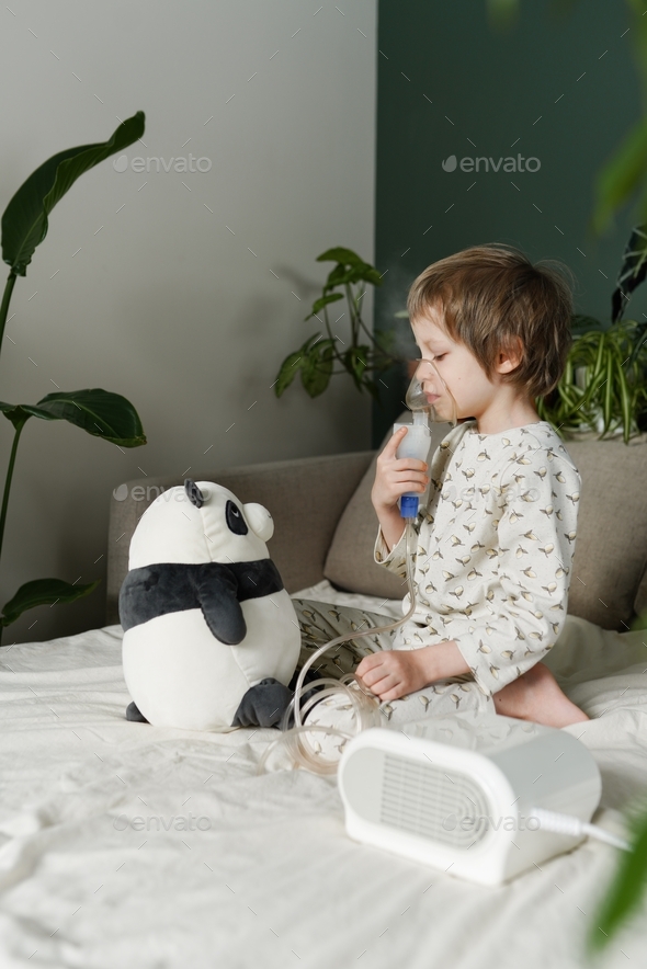 Sick boy playing in bed with his bear toy and nebulizer inhaler. Unwell ...