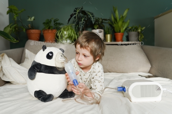 Sick boy laying in bed with his bear toy and nebulizer inhaler. Unwell ...