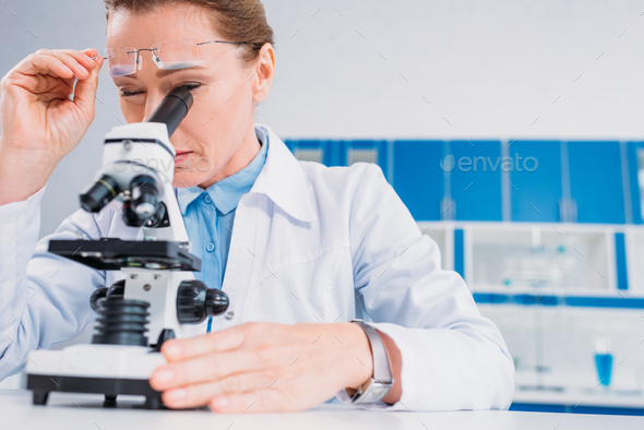 female scientist in lab coat and eyeglasses looking through microscope ...