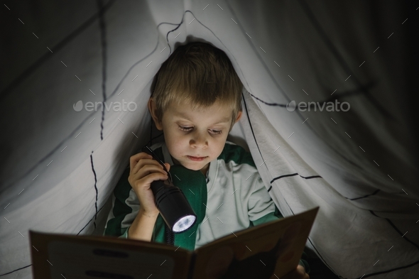 Little boy reading a book at night covered with blanket with lantern ...