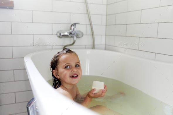 Positive little girl sitting in water of bath with bar soap while ...