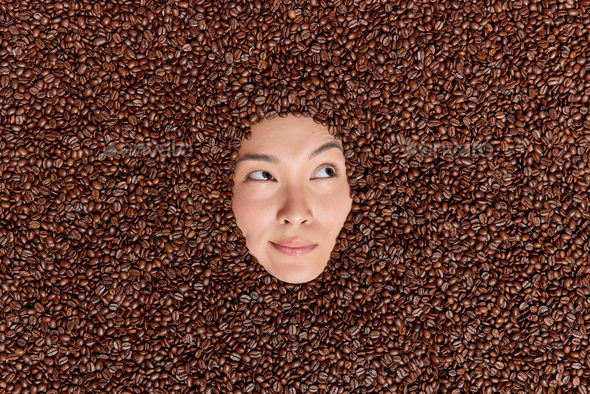 Pensive female model drowned in brown coffee beans looks thoughtfully ...