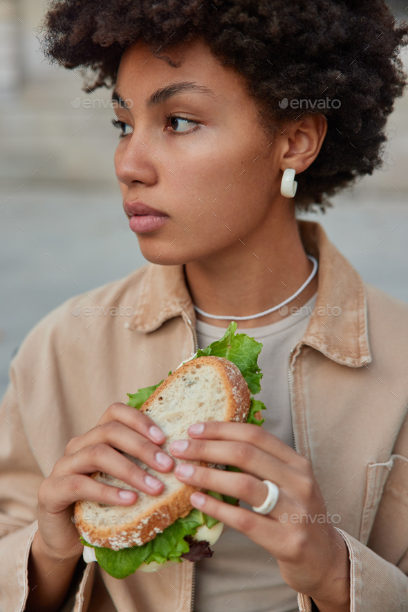 Hungry young woman has snack at street holds delicious sandwich eats ...