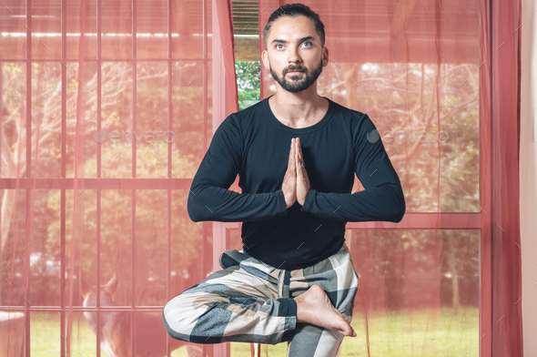 A young man doing tree pose in yoga exercises with his palms together ...