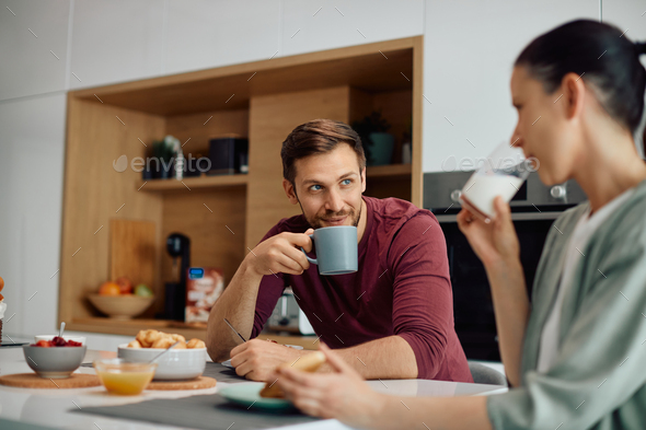 Smiling couple talking while eating breakfast together at dining table ...