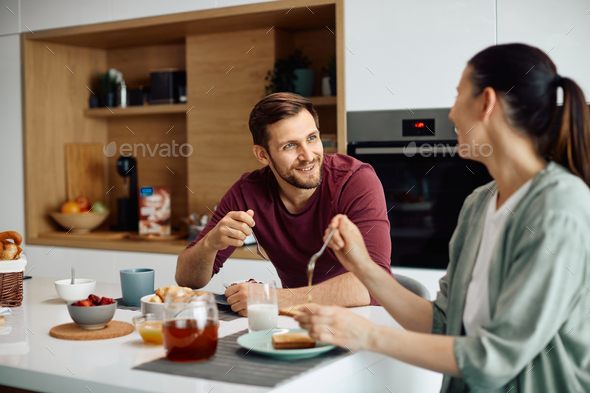 Happy couple talking while eating breakfast at dining table at home ...