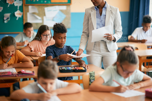 Black elementary student receiving exam paper from teacher during class ...