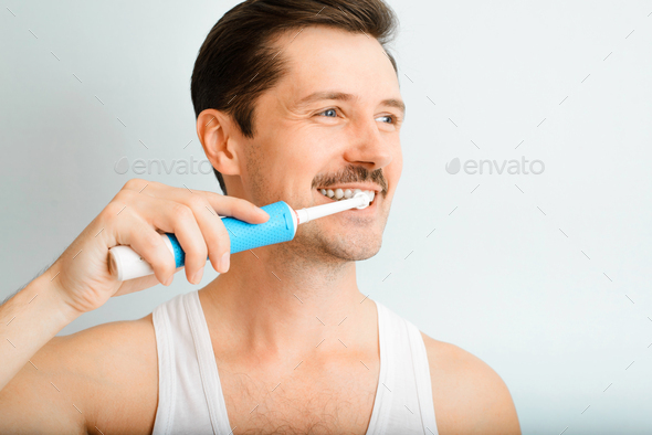 Handsome smiling young man brushing white teeth with modern electric ...