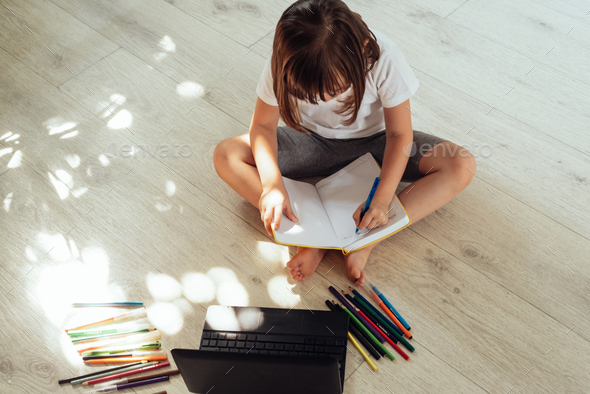 Child sits on the floor with a laptop, pen, notepad and colored pencils ...