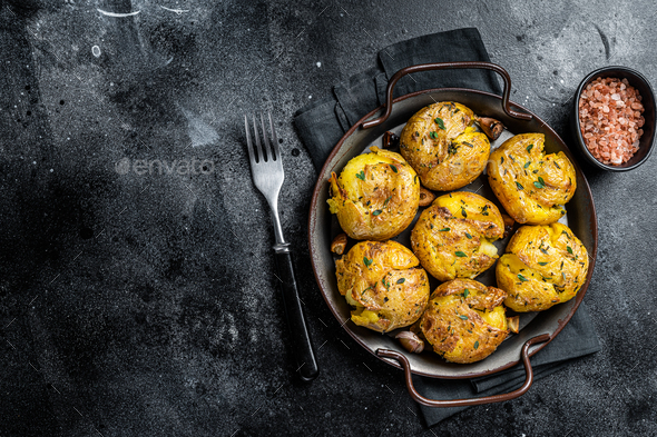 Broken potatoes baked in oil with herbs. Black background. Top view ...