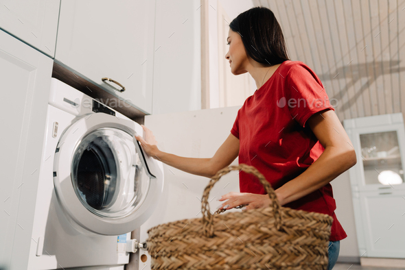 Young woman putting clothes at washing machine while doing laundry ...