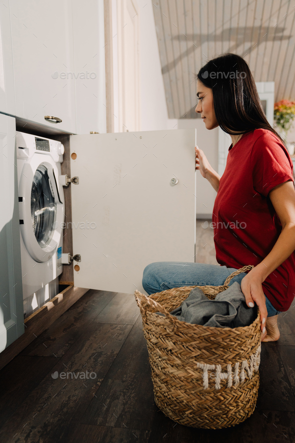 Young woman putting clothes at washing machine while doing laundry ...