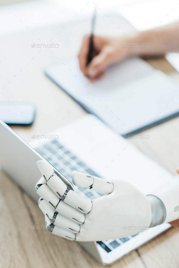 close-up view of robotic arm using laptop and human hand taking notes ...