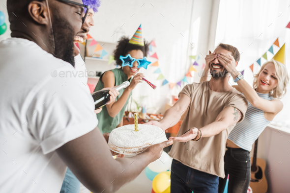 Partying diverse people covering eyes of young friend and greeting him ...