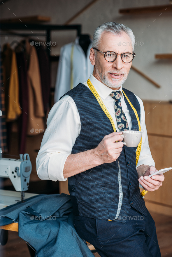 handsome tailor standing with cup of coffee and smartphone at sewing ...