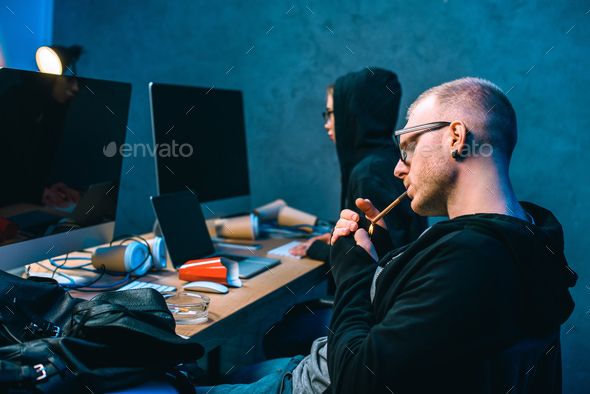 side view of hacker working on malware with accomplice and smoking ...