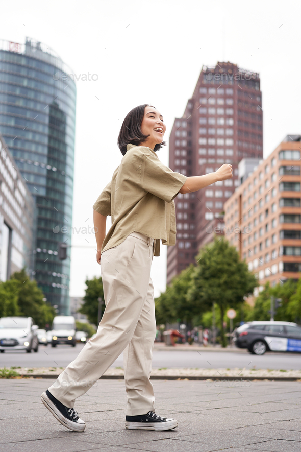Vertical shot of young asian woman posing happy, raising hands up and ...