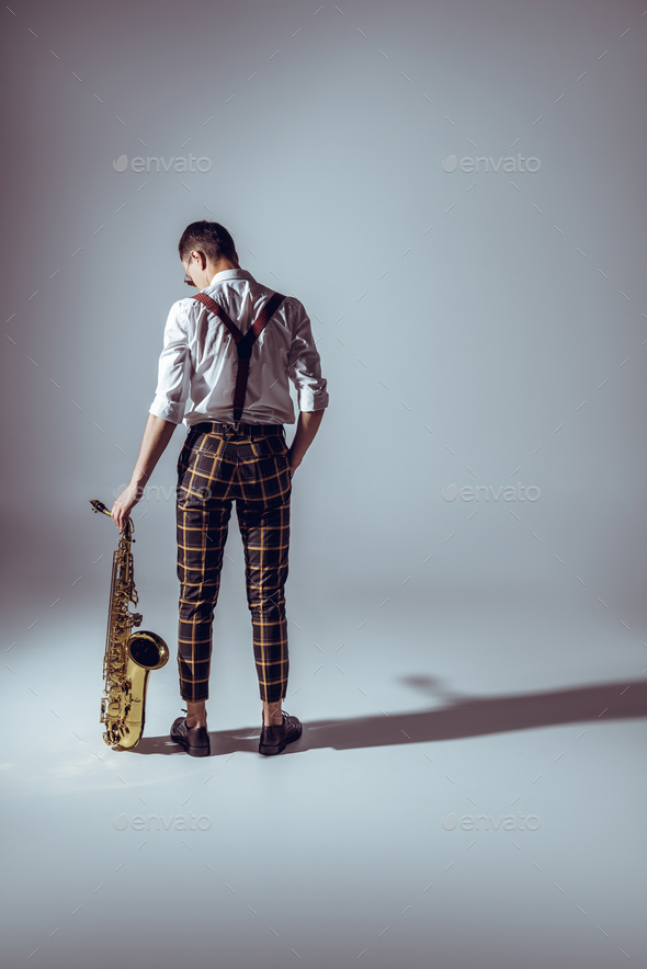 back view of stylish young musician standing with saxophone on grey ...