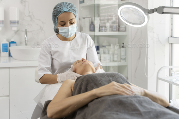 Charming young beautician in white lab coat examining her senior female ...