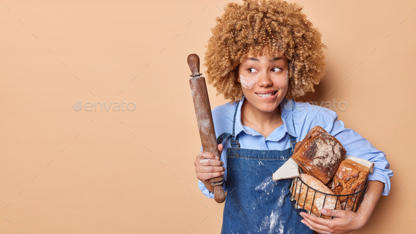 Busy female baker bites lips looks aside holds wooden rolling pin and ...