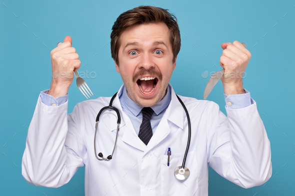 Hungry young male doctor holding fork and knife on hand ready to eat ...