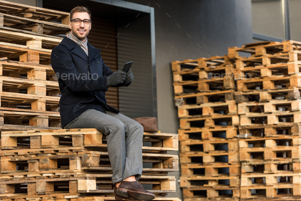 handsome smiling man using smartphone and sitting on wooden pallets ...