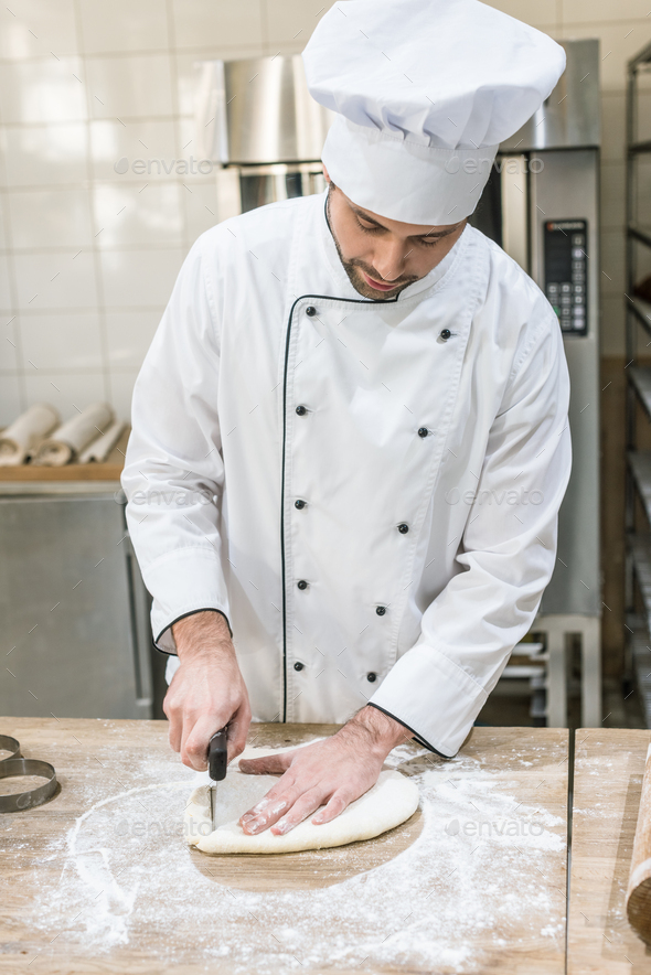 Baker in white chefs uniform cutting dough on wooden table at ...