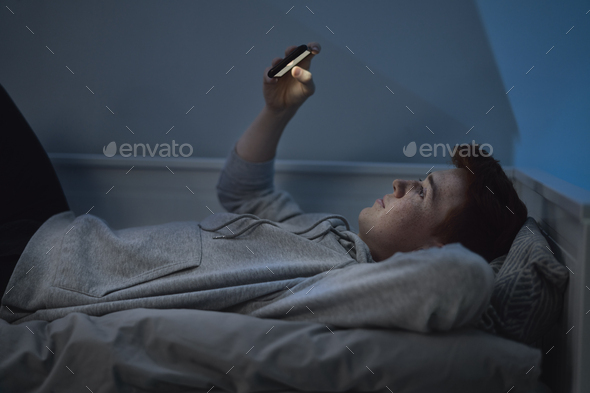 Cheerful caucasian teenage boy watching mobile phone while lying in bed ...