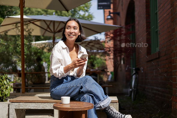 Stylish young asian woman sitting with cup of coffee in an outdoor cafe, smiling and looking ...