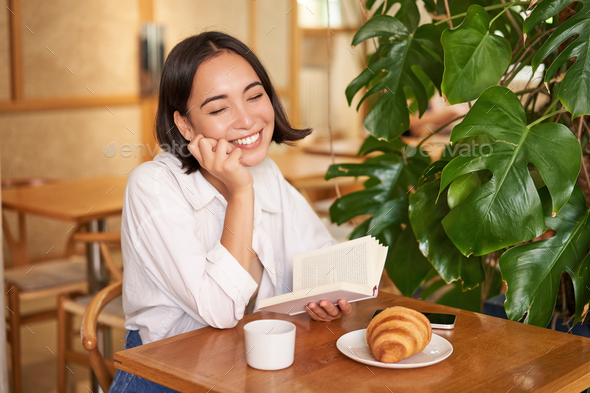 Romantic asian woman sitting with book in cafe, eating croissant and ...