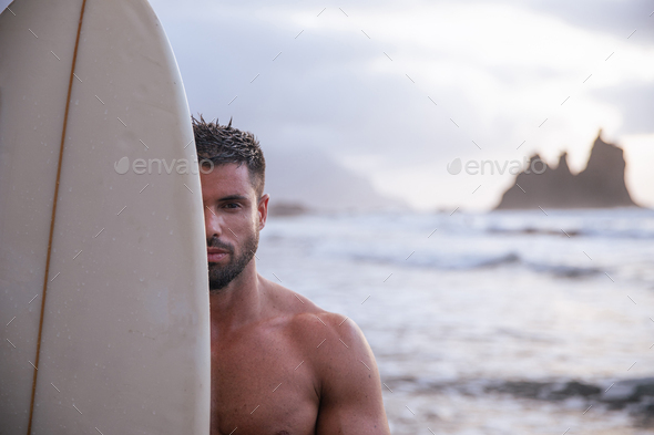 Portrait of a surfer covering half of his face with surfboard, muscular ...