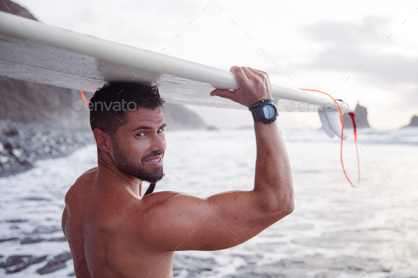 Attractive surfer smiles with his surfboard resting on his head, he is ...