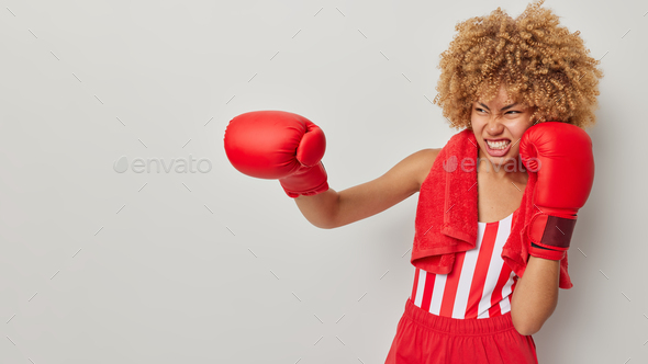 Studio shot of angry curly female boxer ready for fighting makes punch ...