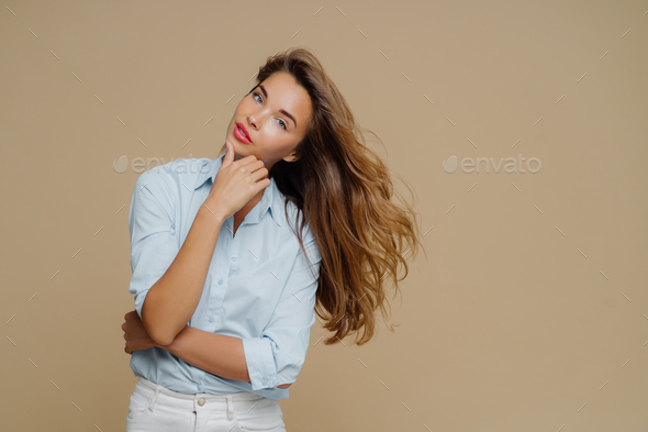 Half length shot of attractive female touches chin, keeps hands partly ...