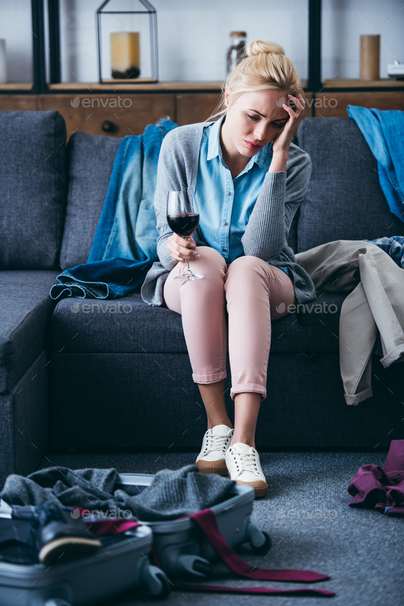 sad woman sitting with glass of red wine while packing in living room ...