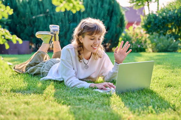 Young female lying outdoor on grass with laptop talking with friends ...