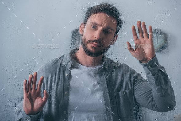 handsome upset man touching window with raindrops and looking away ...
