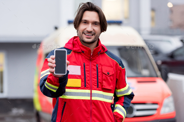 Smiling paramedic in red uniform holding smartphone with blank screen ...