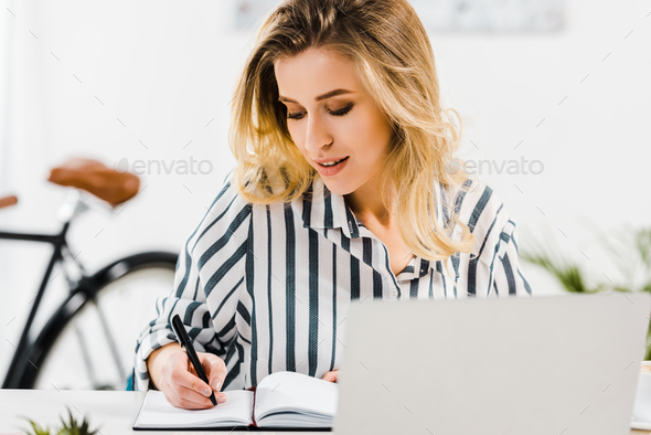 Charming blonde woman in striped shirt writing in notebook at workplace ...