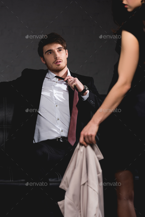 selective focus of handsome man sitting on couch and taking off tie ...