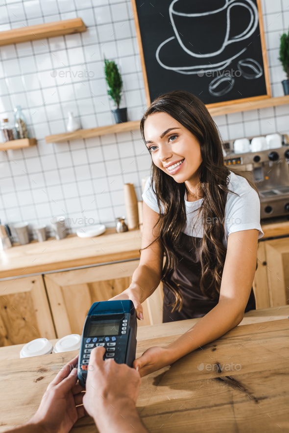 attractive cashier standing near bar counter in brown apron and holding ...