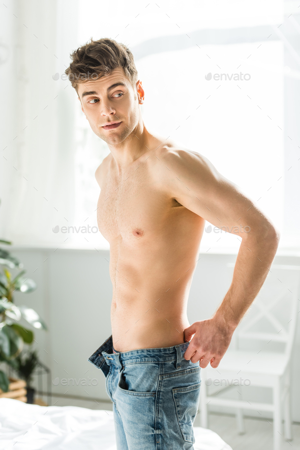 handsome man with bare torso standing near window and putting on blue ...