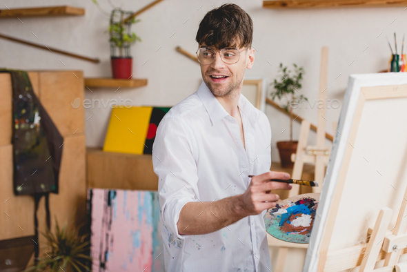 selective focus of smiling artist in white shirt and glasses painting ...