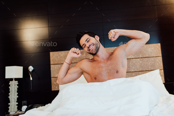 cheerful muscular man stretching in bed after wake up Stock Photo by LightFieldStudios
