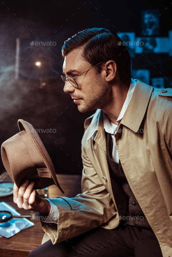 Pensive detective in glasses and trench coat sitting on table and ...