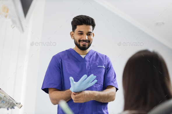 Confident bearded male dentist puts on blue rubber gloves, standing in a dental office Stock ...