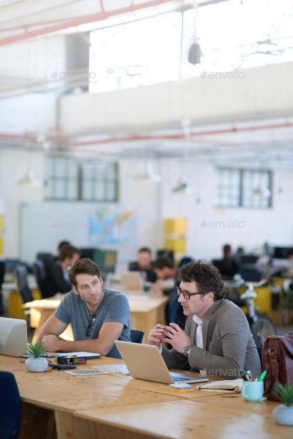 Shot of two colleagues talking together while working on laptops at a ...