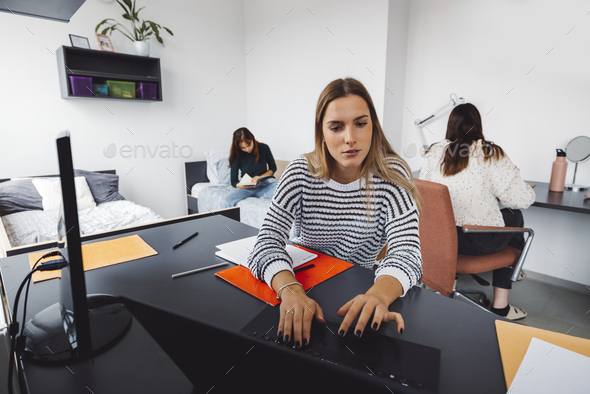 Female student doing homework on her laptop while her roommates are ...