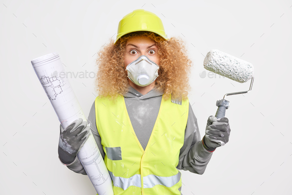 Surprised female architect looks surprisingly holds paper blueprint and painting roller wears ...