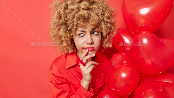 Photo of displeased curly woman has leaked makeup after crying holds heart balloons feels sullen ...